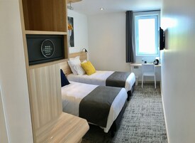 Quarto de hotel com duas camas, uma escrivaninha, uma cadeira, uma televisão e uma janela com cortinas.