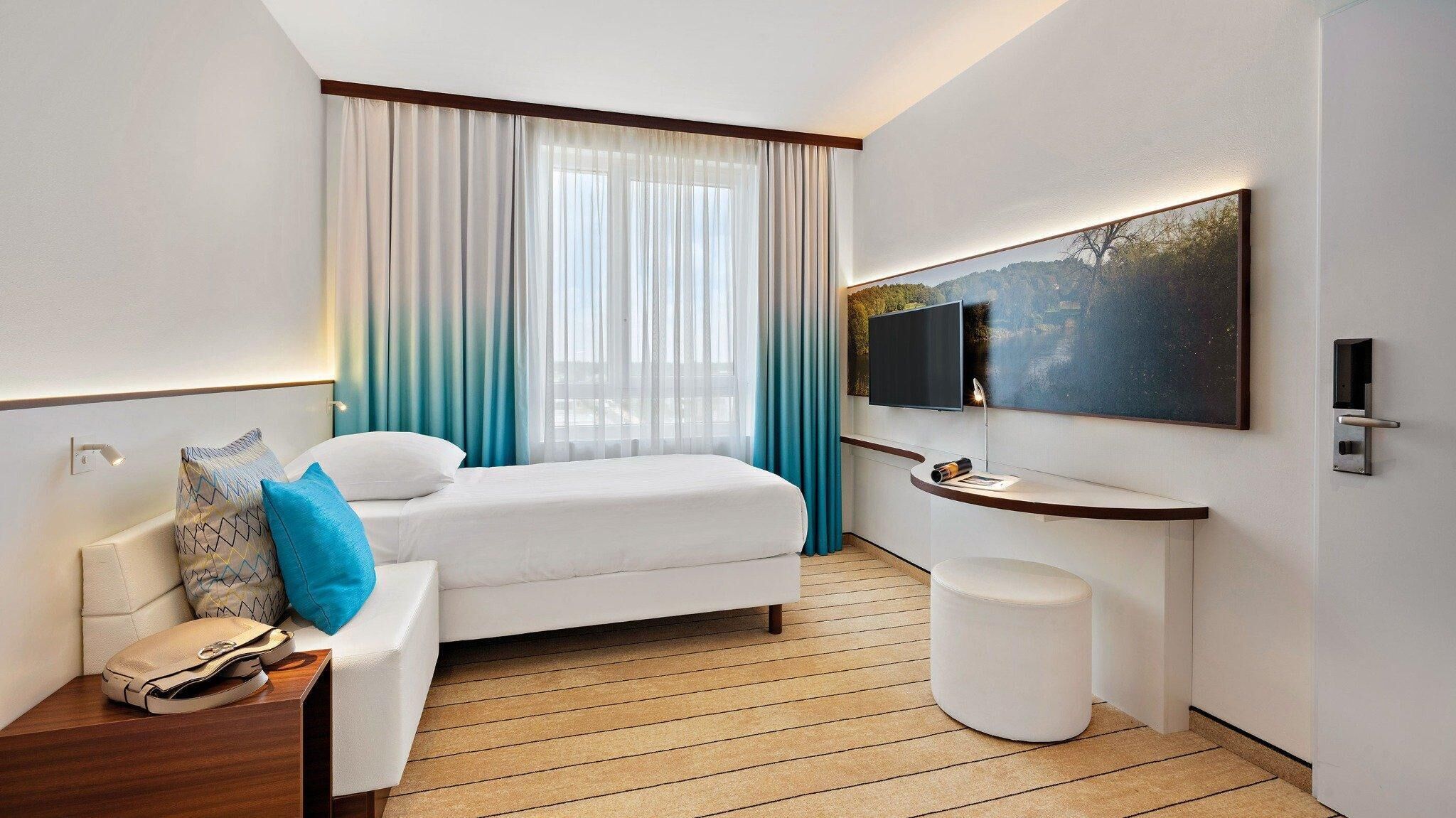 Quarto de hotel moderno com uma cama, mesa de cabeceira, TV e um pequeno banquinho branco.