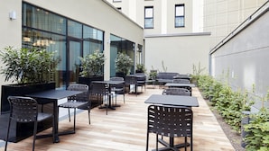 Uma área de pátio ao ar livre com várias mesas e cadeiras, cercada por plantas em vasos e um edifício com grandes janelas.
