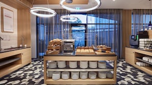 Uma área de café da manhã moderna, com um balcão central exibindo pães e doces, cercada por prateleiras de pratos e um piso estampado.