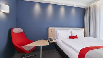 Quarto de hotel com cama, uma cadeira vermelha, uma mesinha e um criado-mudo com telefone.