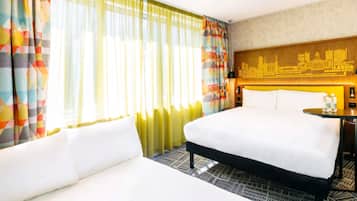 Quarto de hotel com cama, sofá, mesa e garrafas de água, e um painel com vista para a cidade.
