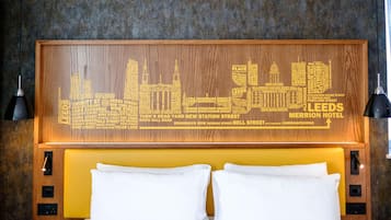 Quarto de hotel com arte de parede retratando uma paisagem urbana, cama com roupa de cama branca e cabeceira de madeira.