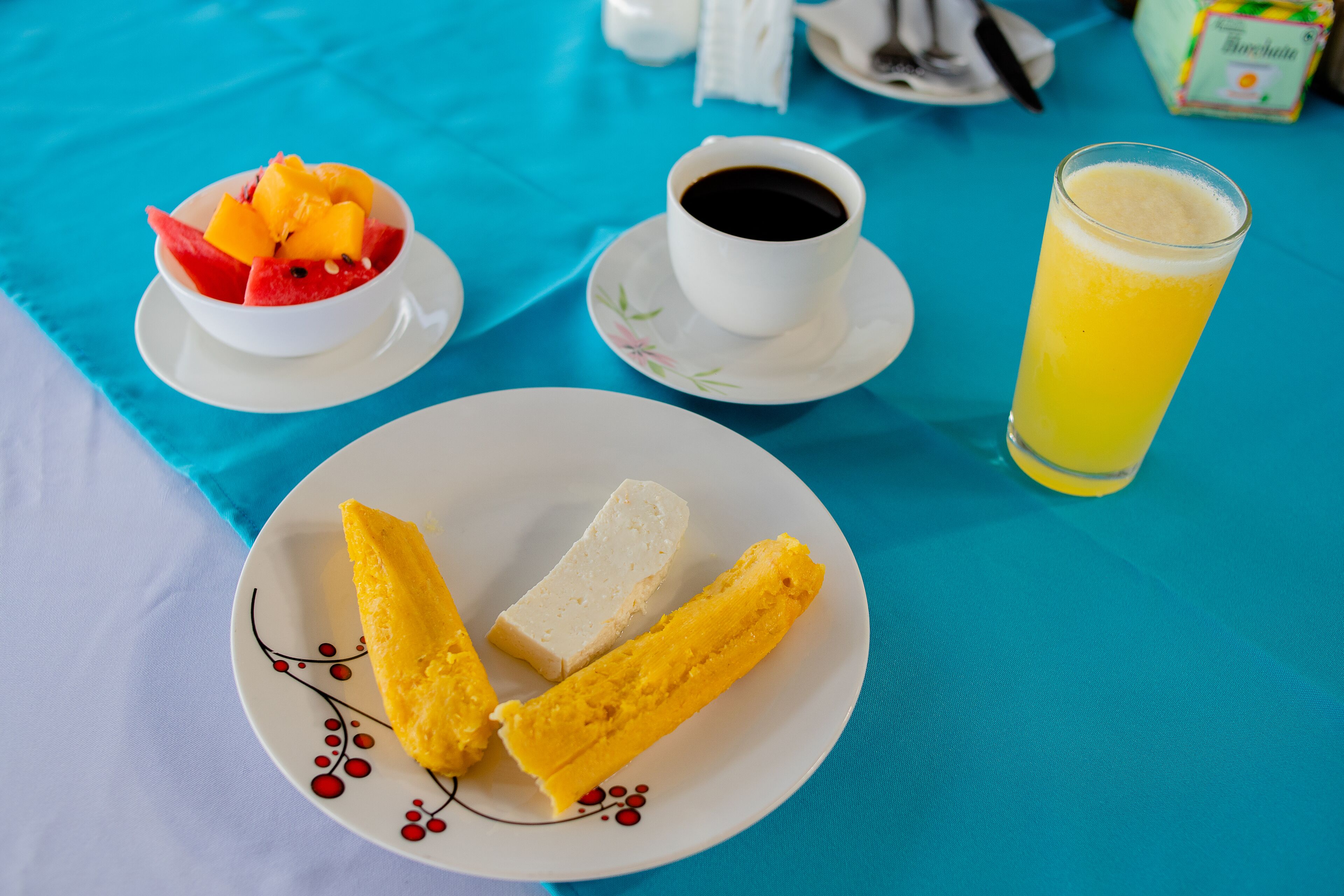 Uma mesa com toalha azul, um prato com duas fatias de pão, uma tigela de fruta, uma xícara de café e um copo de suco de laranja.