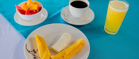 Uma mesa com toalha azul, um prato com duas fatias de pão, uma tigela de fruta, uma xícara de café e um copo de suco de laranja.