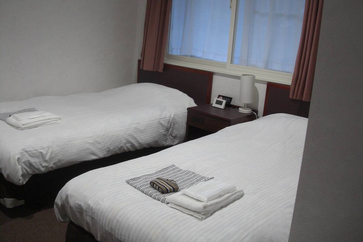 Duas camas de solteiro com lençóis brancos, uma mesa de cabeceira com abajur e uma janela com cortinas.