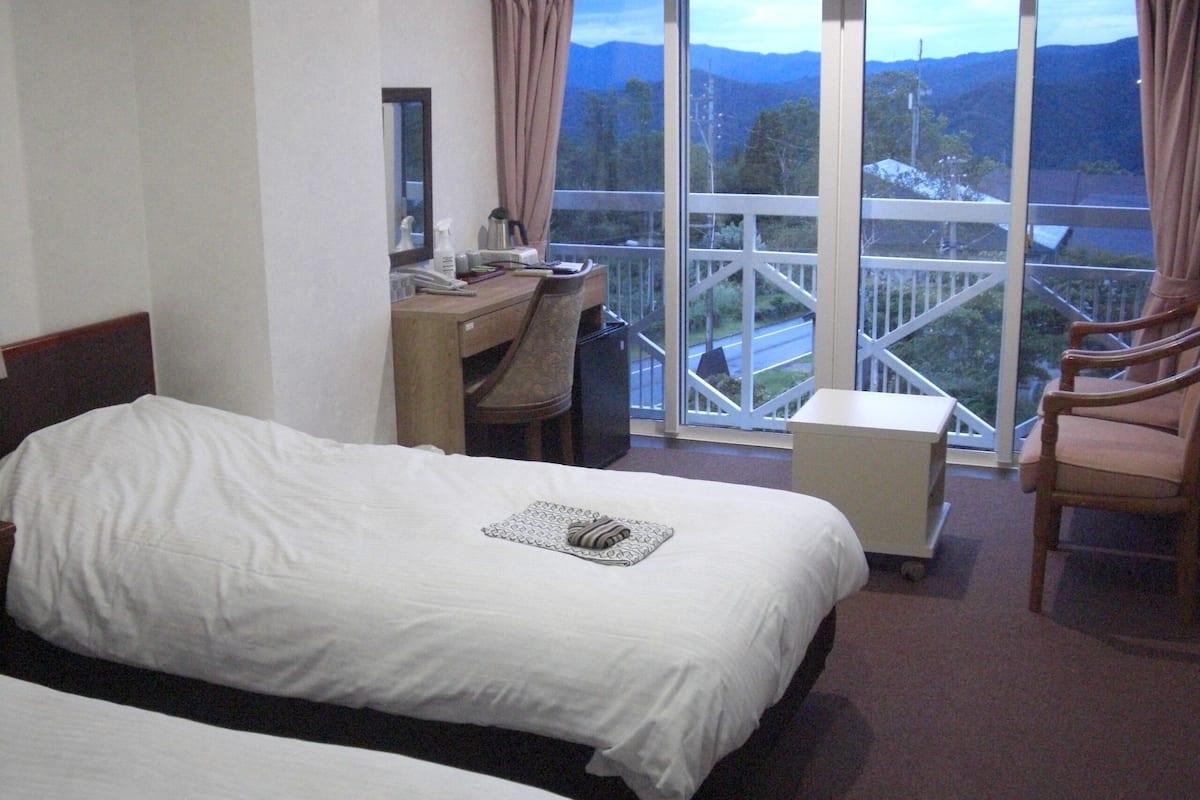 Quarto de hotel com duas camas, uma escrivaninha, uma cadeira e vista para as montanhas através de amplas janelas.