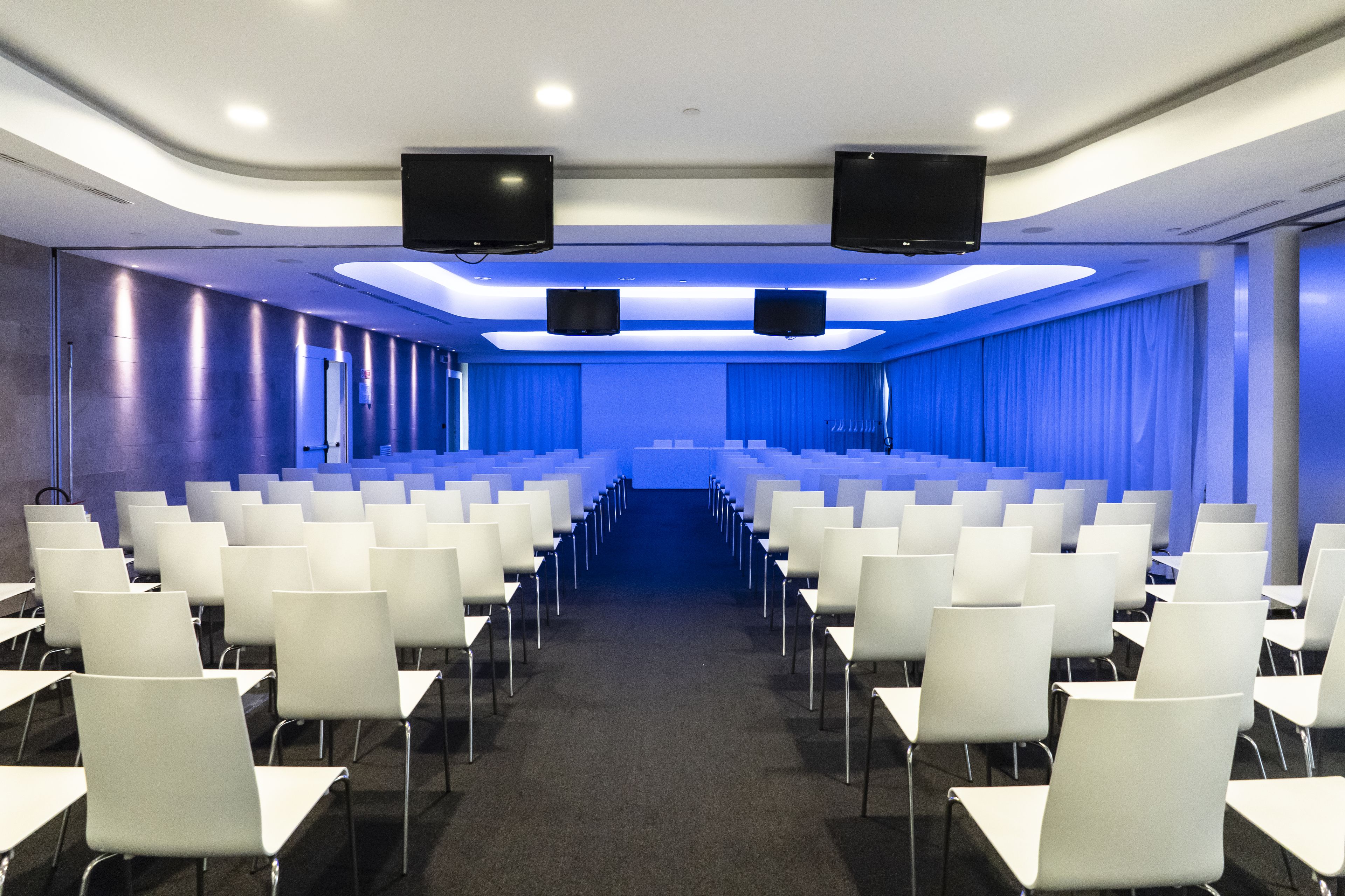 Uma sala de conferências com fileiras de cadeiras brancas, um palco na frente e duas telas fixadas no teto.