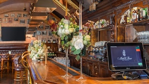 Uma área de bar com balcão de madeira, dois copos altos com flores, uma tela digital exibindo 'Hotel U France Prague' e um bar abastecido com diversas garrafas.