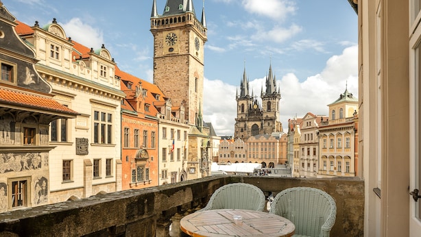 Varanda com cadeiras de vime e uma mesa, com vista para edifícios históricos e um campanário.