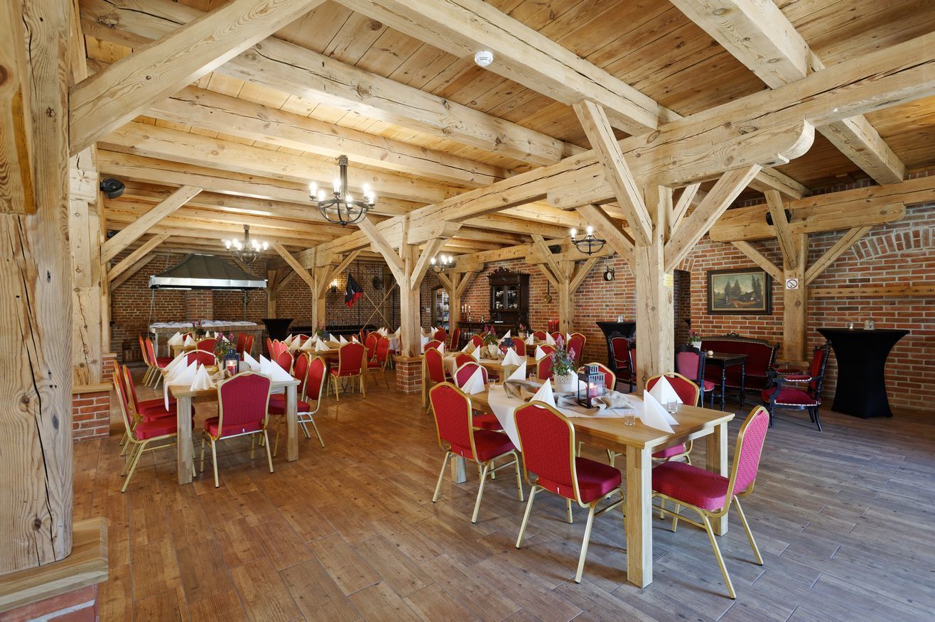 Uma sala de jantar ampla com vigas de madeira e paredes de tijolo, com cadeiras estofadas vermelhas e mesas de madeira.