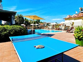 Piscina com mesa de pingue-pongue azul e cadeiras, rodeada por espreguiçadeiras e guarda-sóis.