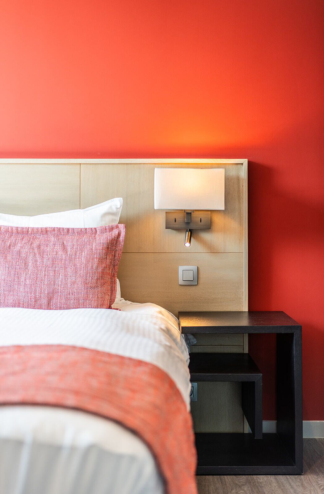 Cama bem arrumada com manta vermelha e branca, travesseiro branco e travesseiro vermelho texturizado. Tem uma mesa de cabeceira com abajur e tomada. A parede é pintada de vermelho.