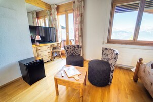 Quarto compacto com piso de madeira, uma pequena geladeira preta, uma mesa de madeira com tábua de corte, duas cadeiras estofadas, uma televisão e uma janela com vista para montanhas.