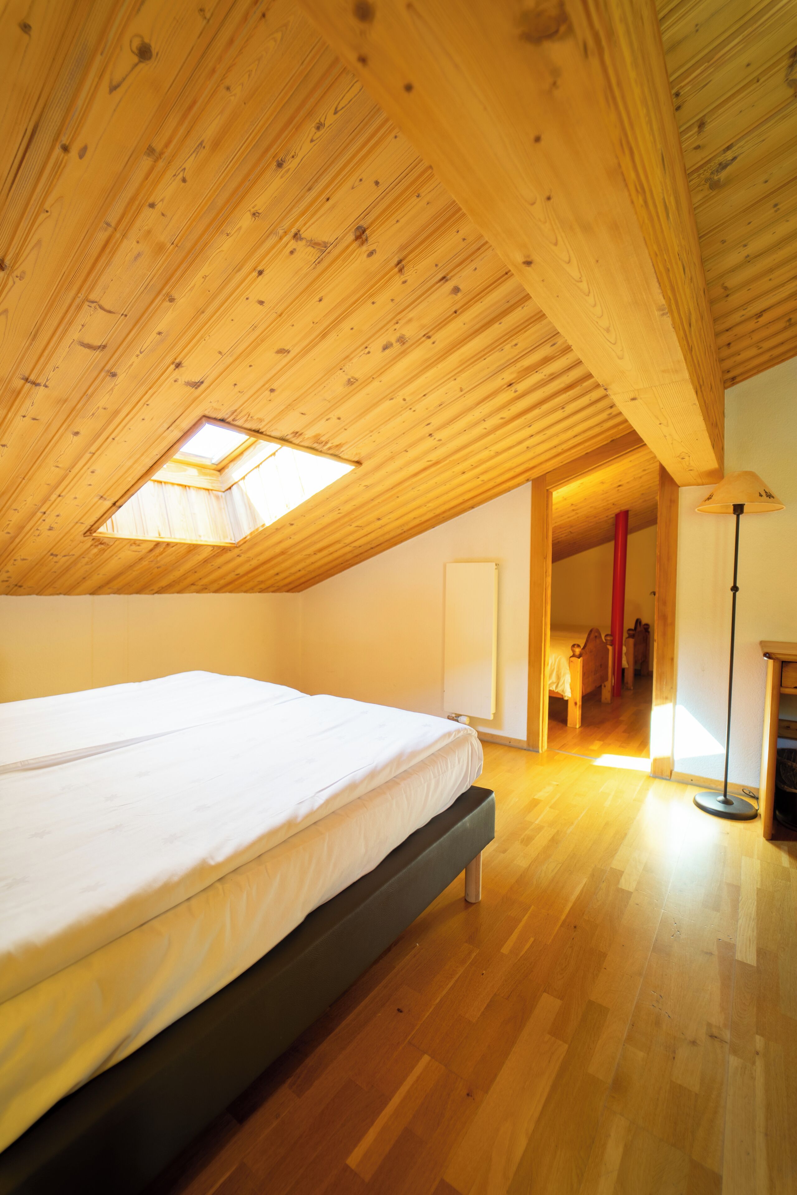 Quarto com teto inclinado, claraboia, cama, luminária de chão e uma porta que dá para outro cômodo.