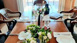Uma mesa de jantar posta com pratos, copos, um jarro de suco de laranja, uma tigela de frutas e um vaso de flores.