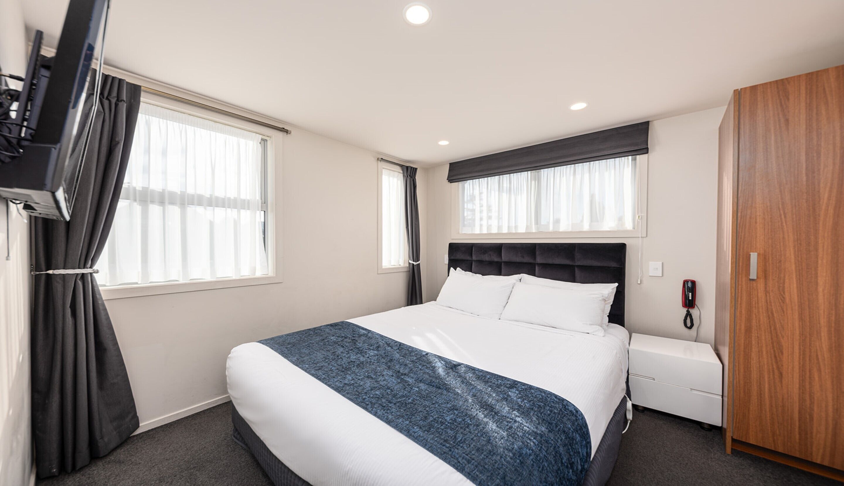 Quarto de hotel com cama, mesa de cabeceira, guarda-roupa de madeira e televisão.