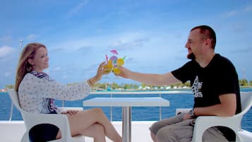 Duas pessoas brindando com bebidas em um barco, com vista para o oceano e ilhas ao longe.