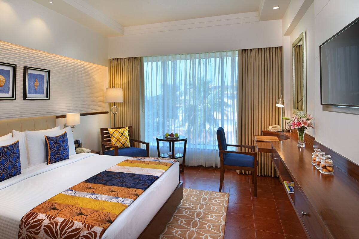 Quarto de hotel com cama, escrivaninha, cadeira, televisão e janela com cortinas.