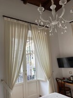 Quarto com porta branca, um candelabro, mesa com televisão e vista para prédios.