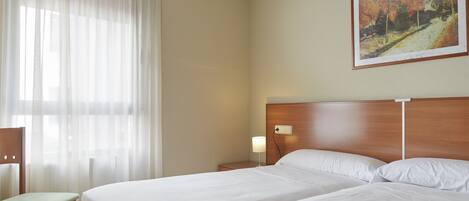 Quarto de hotel com duas camas, uma cadeira, uma mesinha de cabeceira e um quadro na parede.