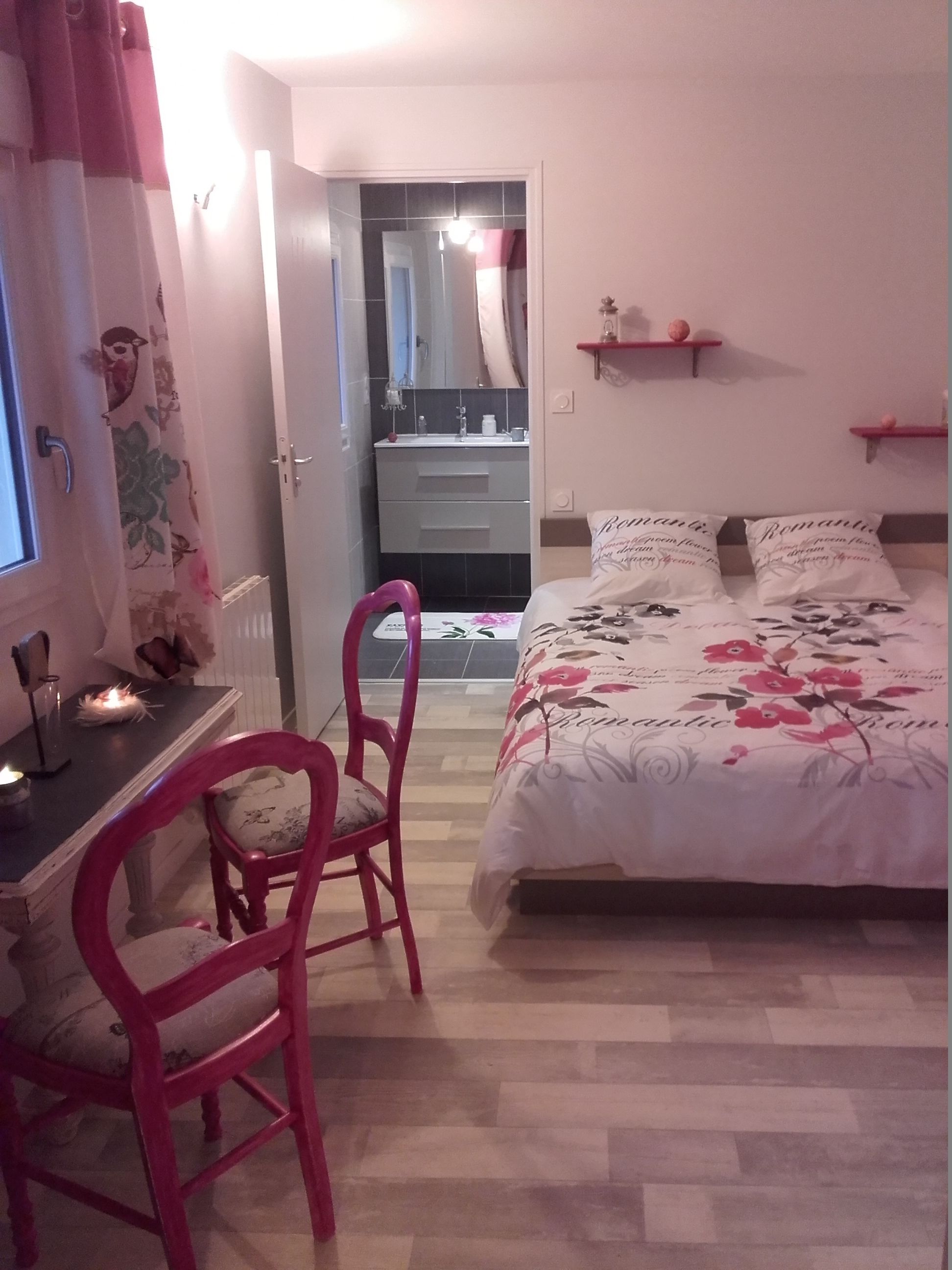 Quarto com cama, escrivaninha, cadeira, espelho e banheiro visível por uma porta aberta.