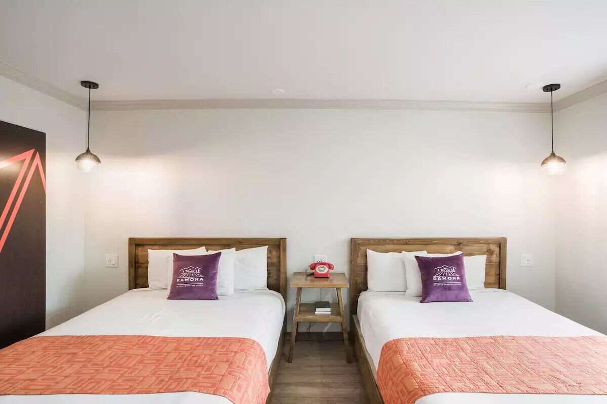 Quarto de hotel com duas camas, cada uma com cabeceira de madeira, roupa de cama branca e mantas com padrão laranja. Pequena mesa de cabeceira com telefone vermelho e um livro. O quarto tem um design simples e clean, com iluminação pendente.