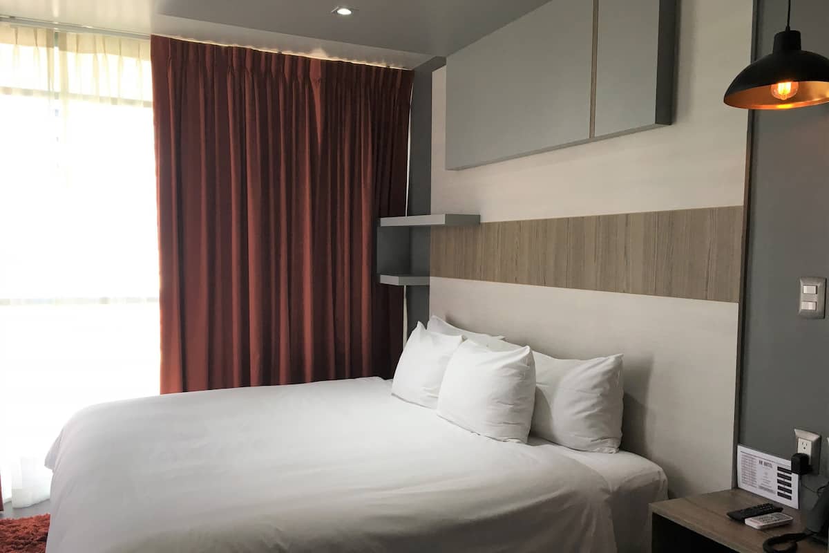 Quarto de hotel com uma cama grande, uma mesinha de cabeceira com telefone e controle remoto, uma televisão na parede e uma janela com cortinas.