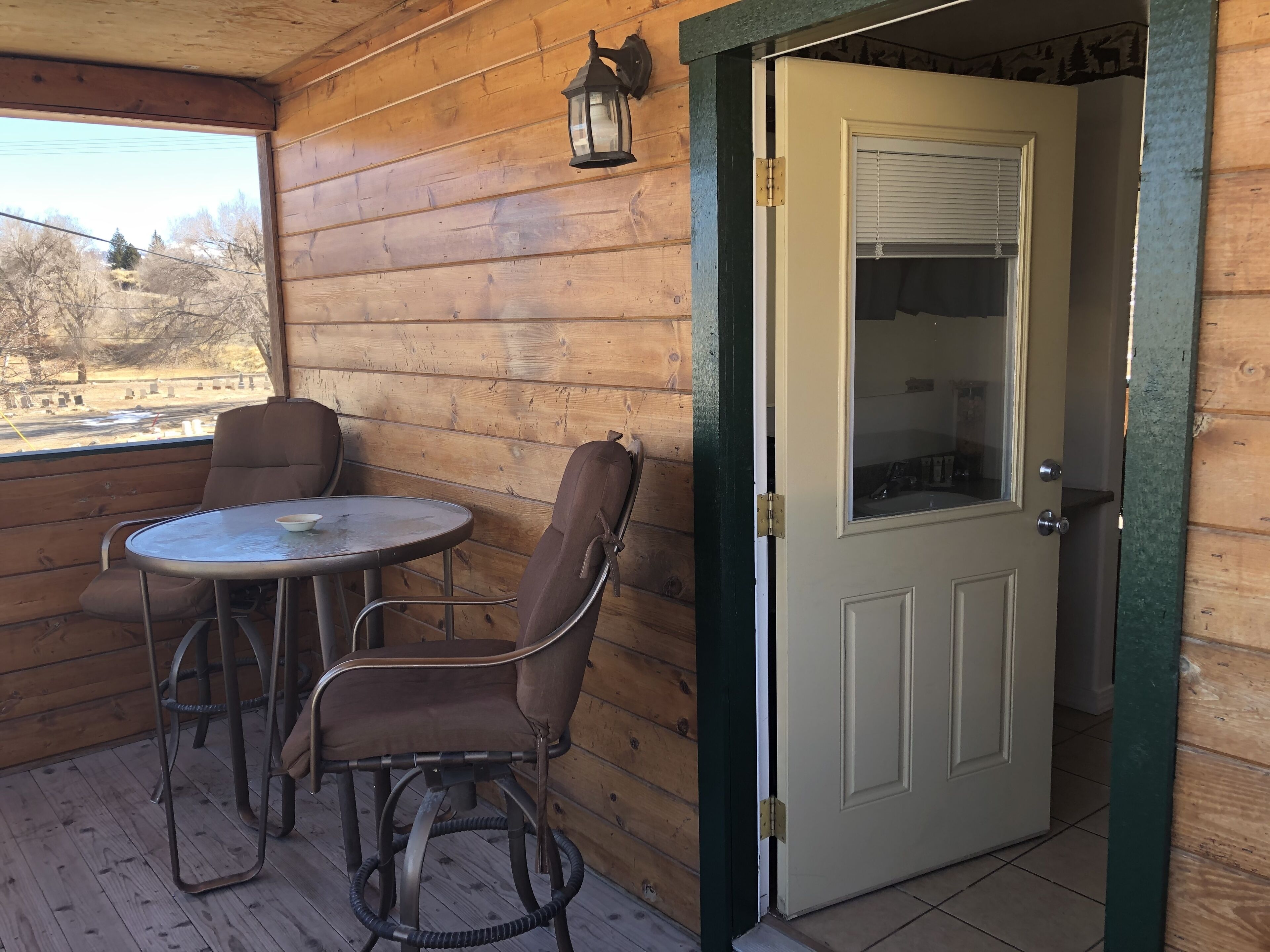 Uma cabana de madeira com uma mesa redonda e duas cadeiras, uma porta com janela, e vista para um cemitério.
