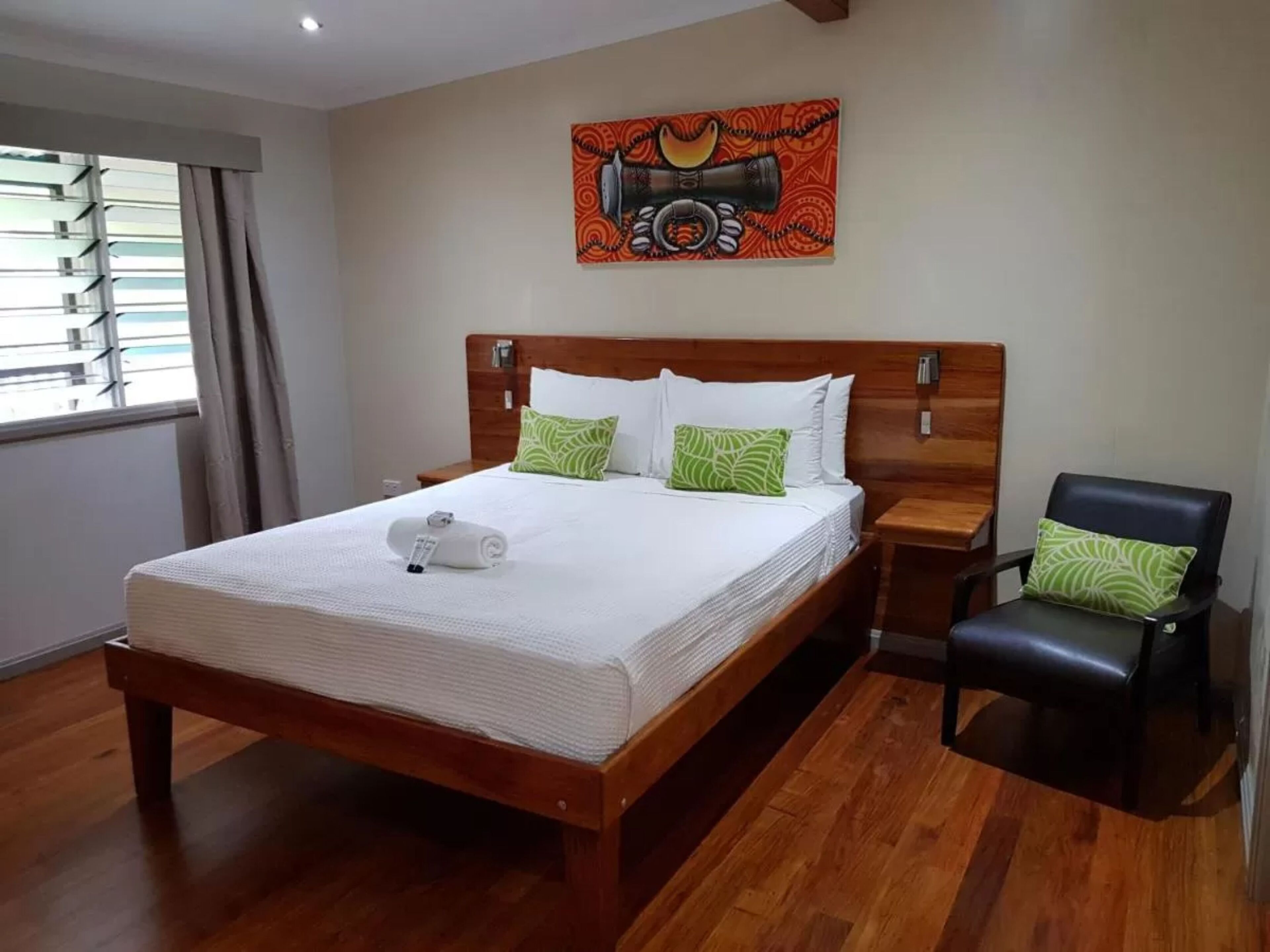 Uma cama bem arrumada com lençóis brancos e almofadas verdes, cabeceira de madeira, mesa de cabeceira de madeira e uma cadeira de couro preto.