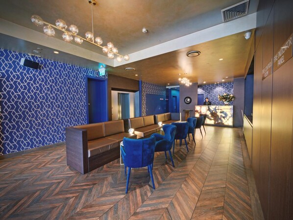 Um lobby de hotel moderno com piso de madeira em padrão herringbone, paredes com padrão azul, um balcão de recepção e áreas de estar.