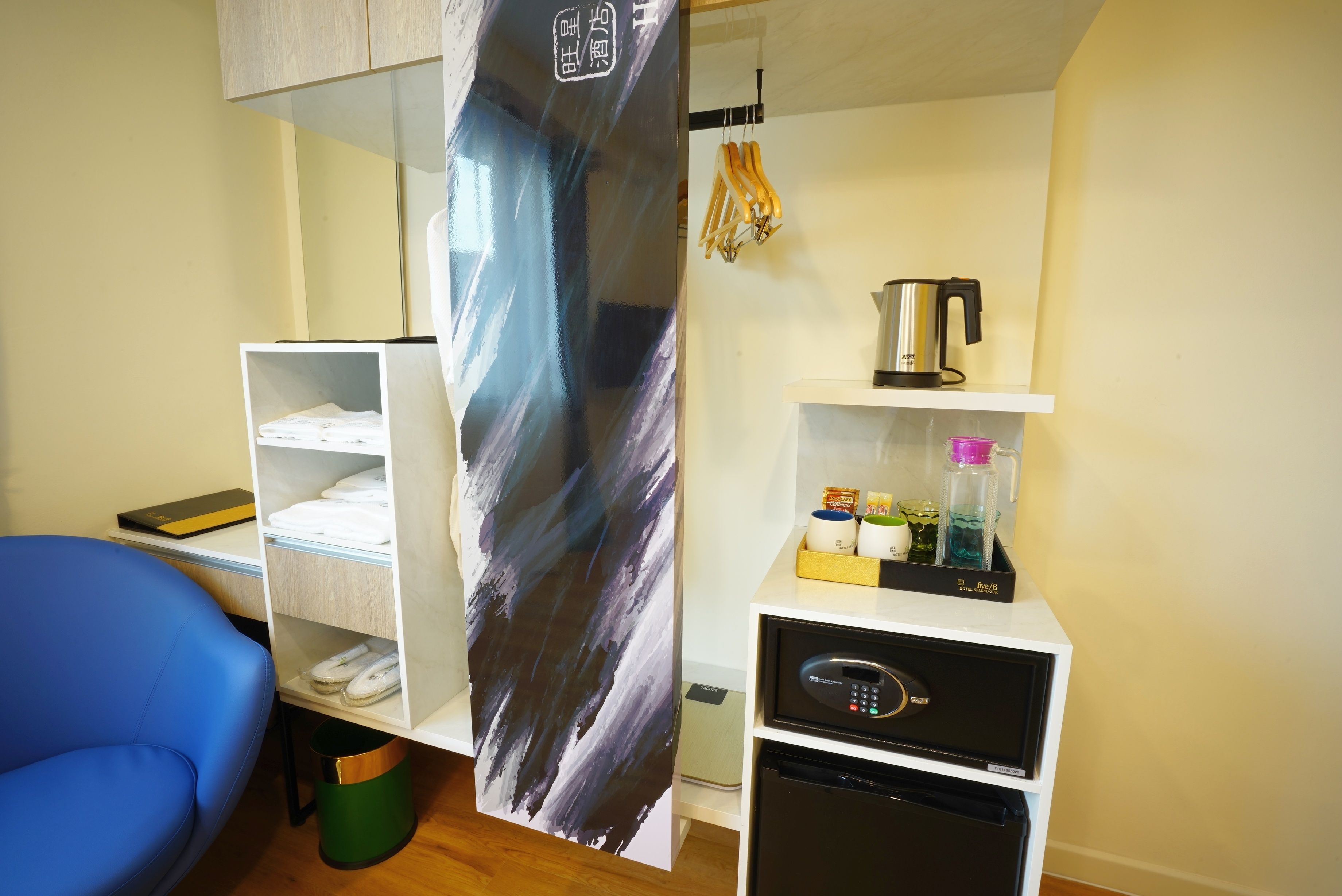 Quarto de hotel compacto com uma cadeira azul, frigobar, cafeteira e um cesto de lixo.