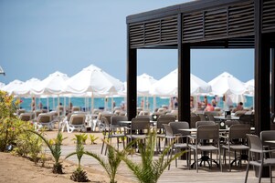 Um restaurante à beira-mar com mesas ao ar livre, guarda-sóis brancos e vista para a praia e o oceano.