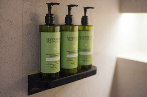 Três frascos de shampoo e condicionador com aroma de verbena limão Seven Plus sobre uma prateleira preta em frente a uma parede texturizada.