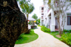 Uma trilha leva a um edifício de fachada branca e varandas, cercado por vegetação e uma árvore.
