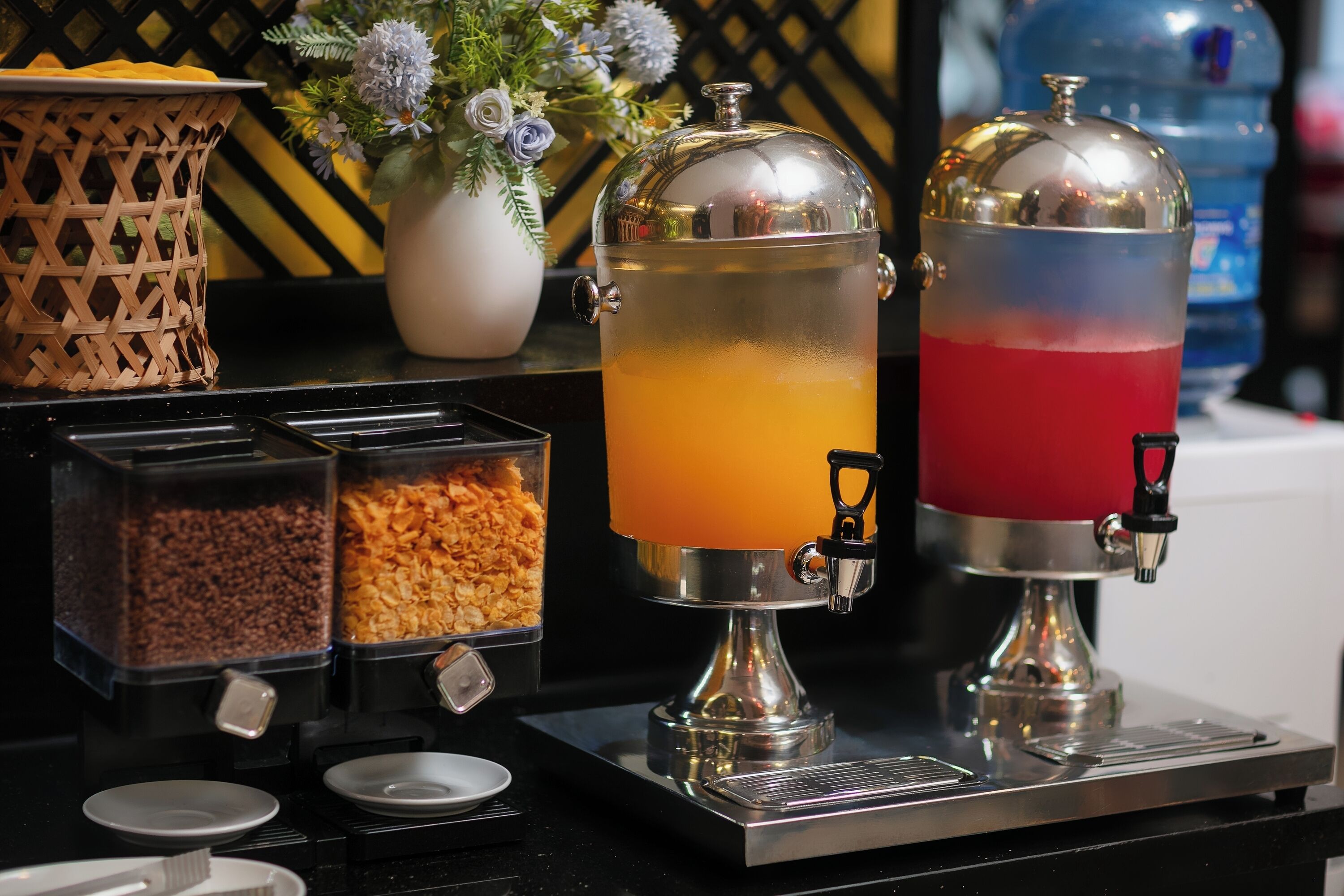 Estação de bebidas com dois dispensadores, um com líquido laranja e outro com líquido vermelho, além de recipientes com granola e cereais.