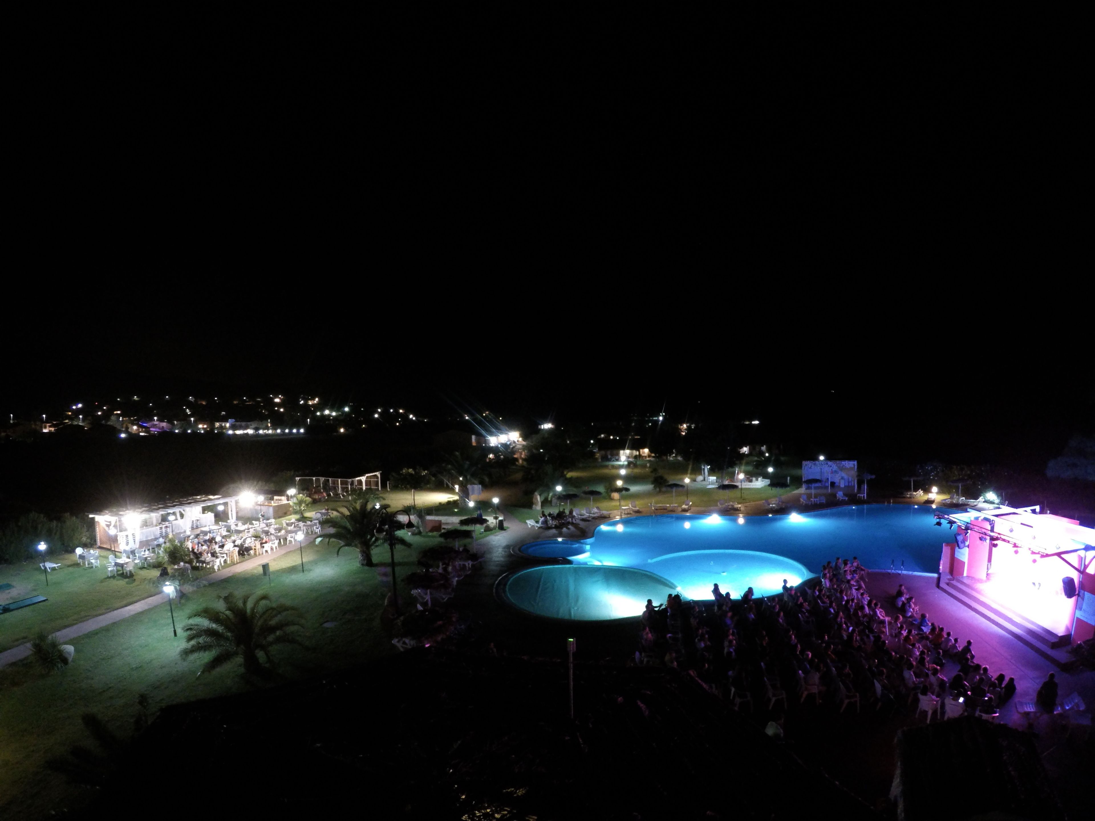 Vista noturna de um resort com várias piscinas, área gramada com assentos e palco para eventos.