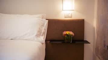Uma cama bem arrumada com lençóis brancos, uma mesa de cabeceira com um vaso de flores e um aplique de parede.