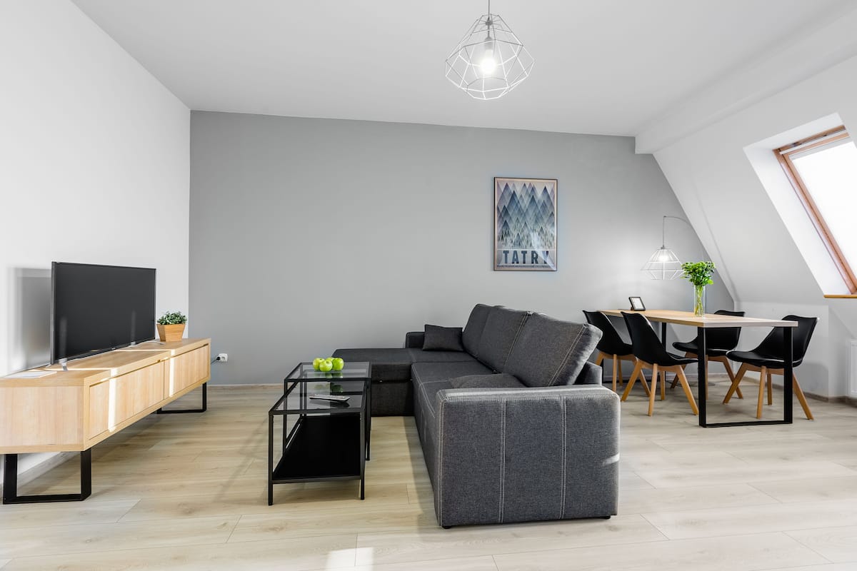 Uma sala de estar moderna com um sofá modular cinza, um armário de madeira com televisão de tela plana, uma mesa de centro de vidro e um espaço de jantar com mesa e cadeiras de madeira.