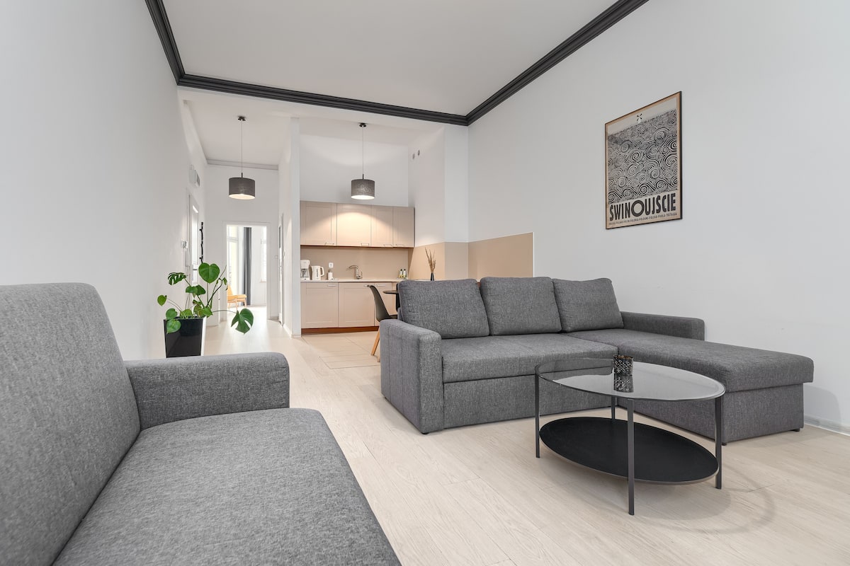 Uma sala de estar moderna com um sofá seccional cinza, uma mesa de centro de vidro e uma cozinha ao fundo.