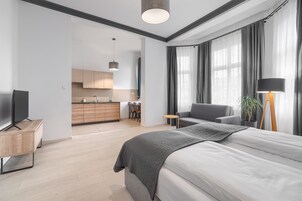 Quarto de hotel moderno com uma cama grande, uma escrivaninha, uma cadeira, uma mesinha e uma televisão. É visível uma área de cozinha ao fundo.