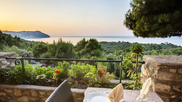 Mesa posta para dois, com pratos, talheres e copos, com vista panorâmica da costa, árvores e montanha ao longe.