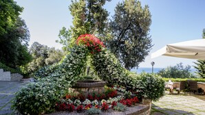 Uma fonte de pedra rodeada por diversas flores e vegetação, com área de estar ao ar livre e vista para o mar.