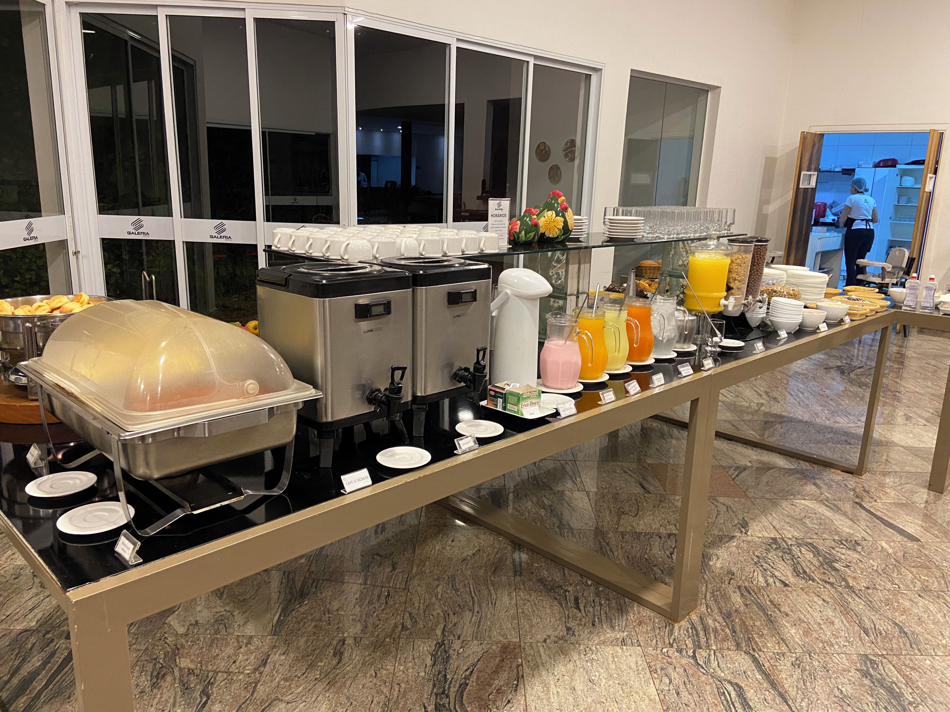 Buffet montado com diversos distribuidores de comida e bebida, um ponto de apoio coberto para alimentos e uma cozinha visível através de portas de vidro.
