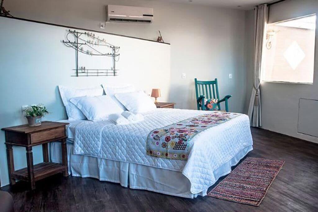 Uma cama bem arrumada com colcha, uma mesa de cabeceira com abajur, uma cadeira e uma janela com cortinas.