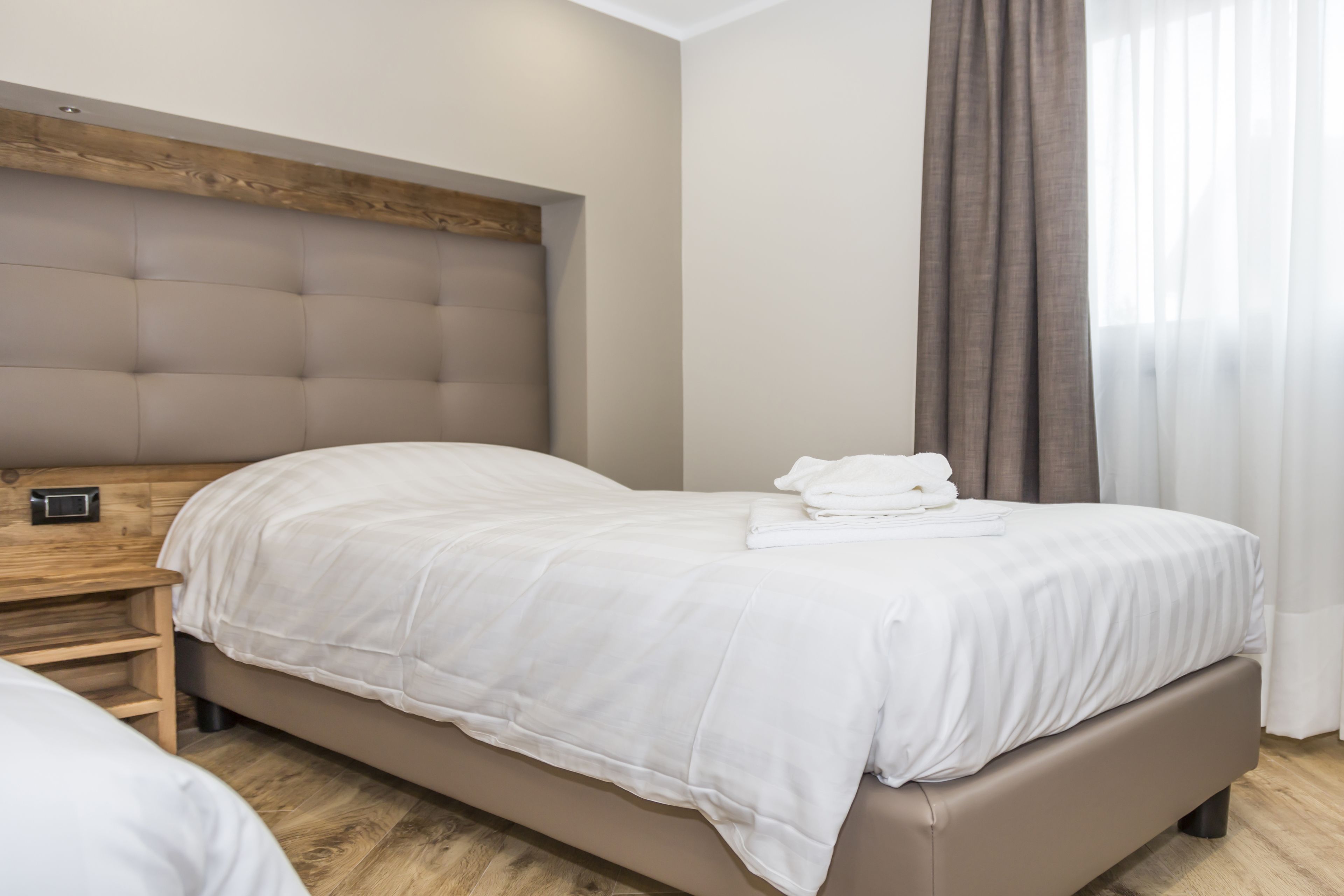 Quarto de hotel moderno com uma cama grande, cabeceira de madeira, mesa de cabeceira com abajur e uma janela com cortinas.
