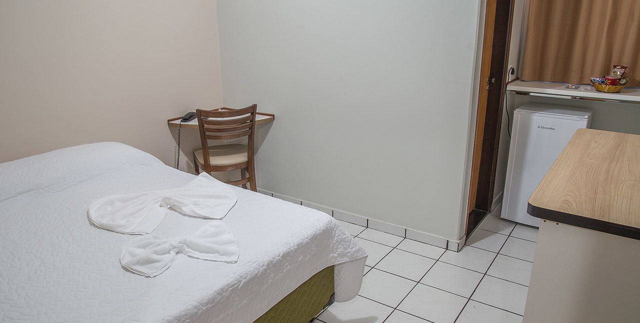 Quarto de hotel pequeno com uma cama, uma cadeira de madeira, uma mesa com telefone e uma área de cozinha compacta.