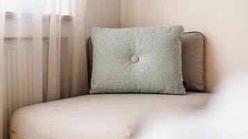 Canto de um quarto com um sofá claro, uma almofada e cortinas leves.