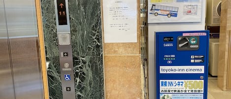 Um elevador com visor digital, uma placa com horário de funcionamento e um anúncio de cinema Toyoko Inn.
