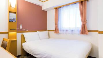 Quarto de hotel com uma cama grande, uma escrivaninha, uma cadeira, uma mesa de cabeceira, um telefone fixado na parede e uma janela com cortinas.
